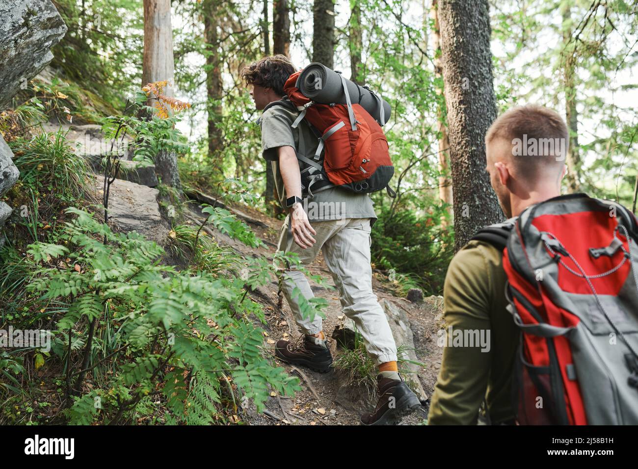 Rear view of young men with backpacks climbing steep cliff while hiking ...