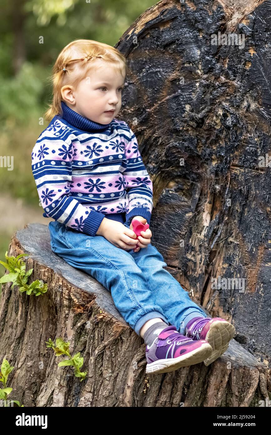 A little girl toddler is sitting on a stump in a spring park. Walk ...