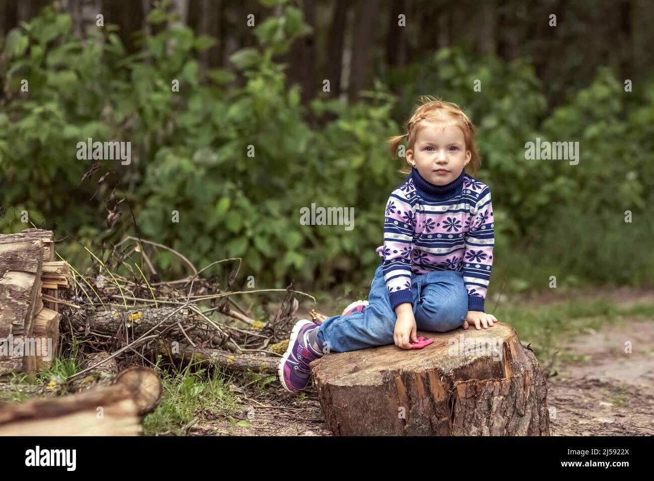 A little girl toddler is sitting on a stump in a spring park. Walk ...