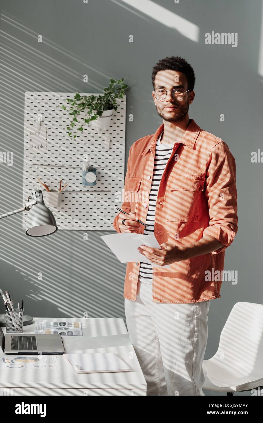 Portrait of content handsome young black manager standing with paper at ...