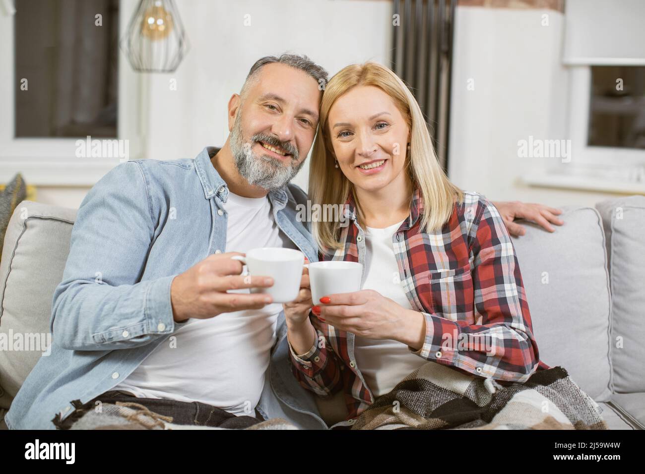 Middle aged couple in casual wear sitting on cozy sofa with cups of ...
