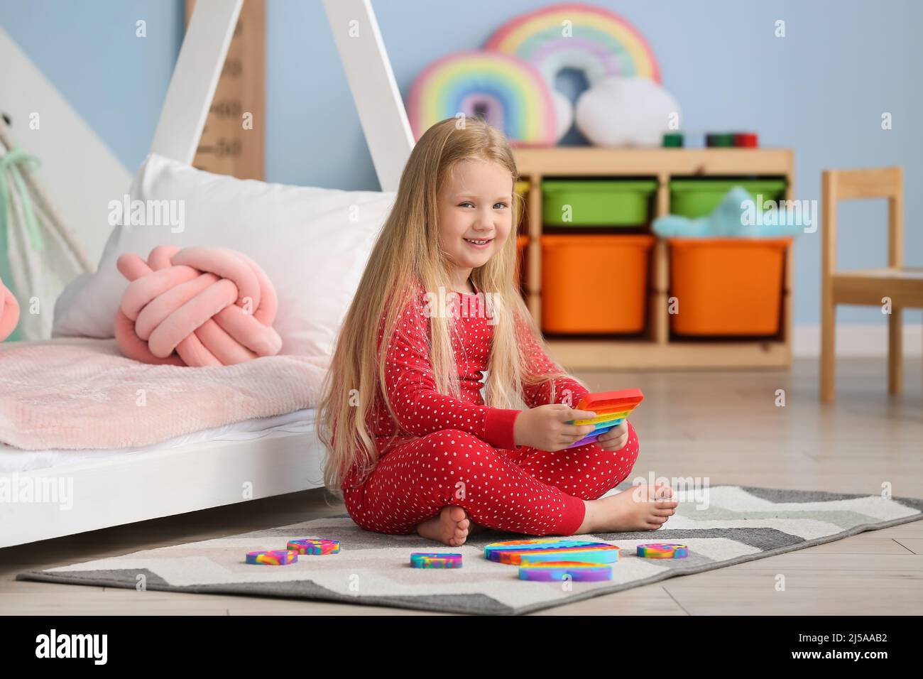 Pretty girl sitting on floor near bed and holding pop it fidget toy in ...