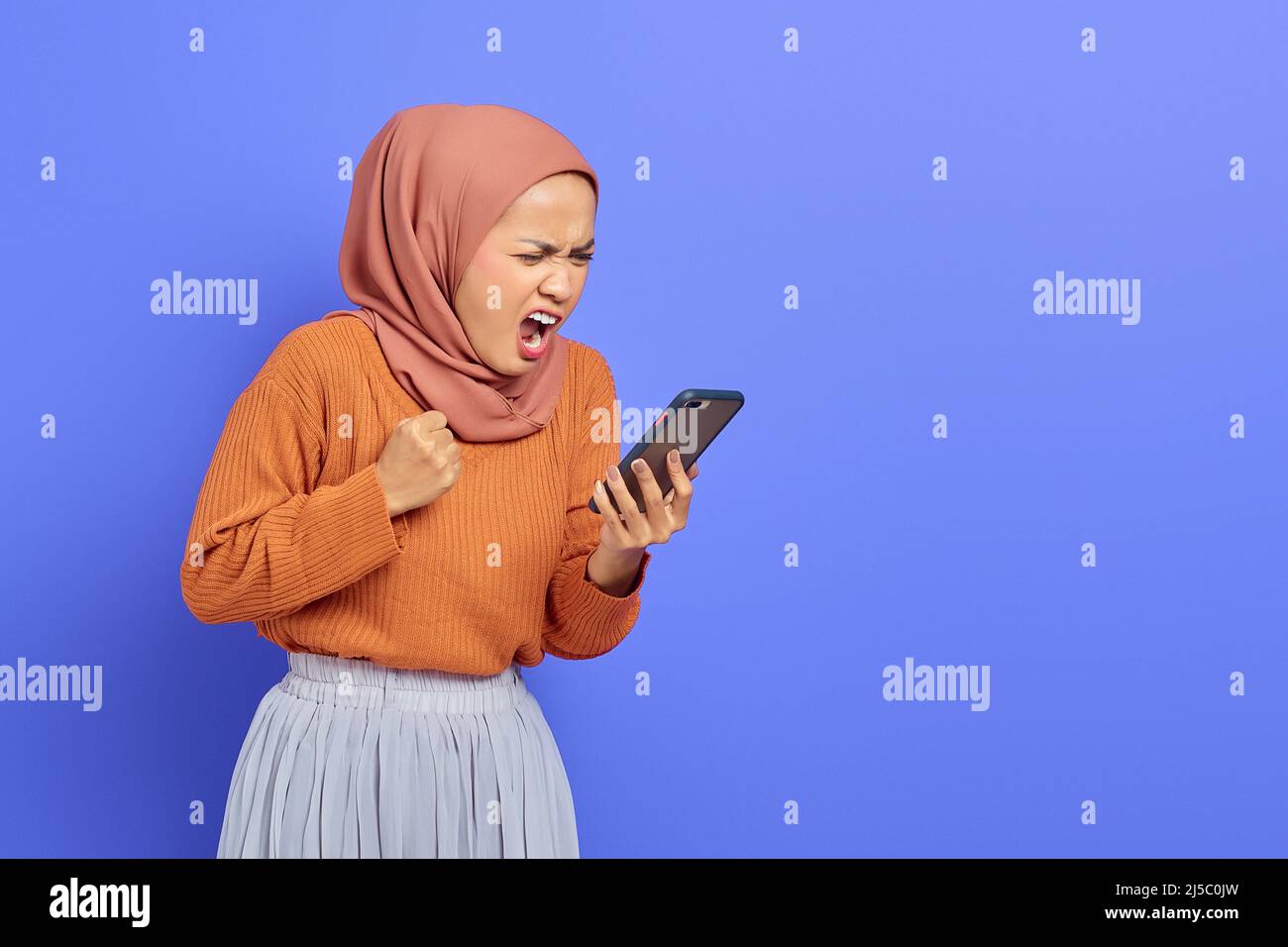 Angry young Asian woman in brown sweater and hijab using smartphone ...