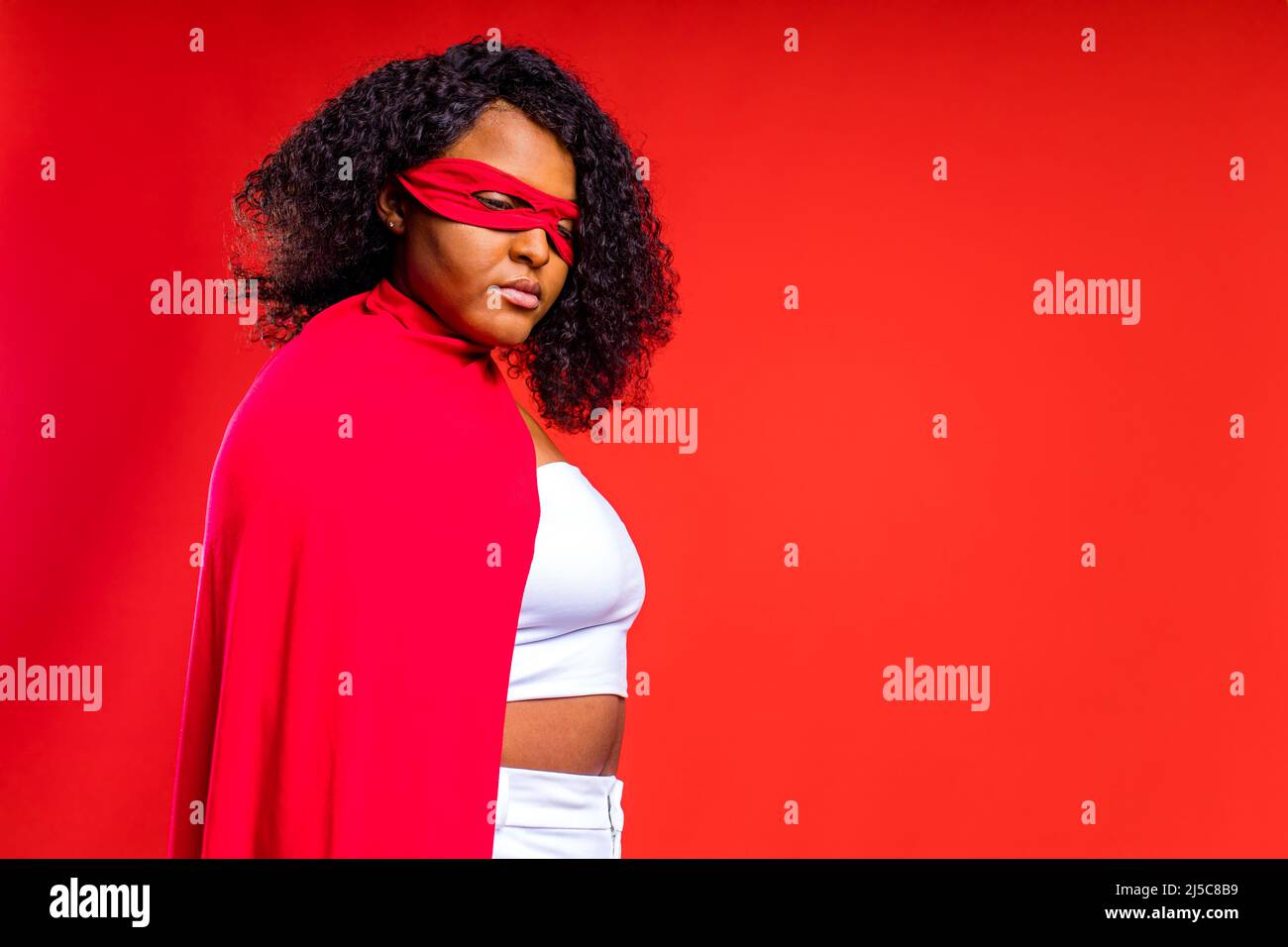 spanish woman in eye mask and superhero cloak in red studio background ...