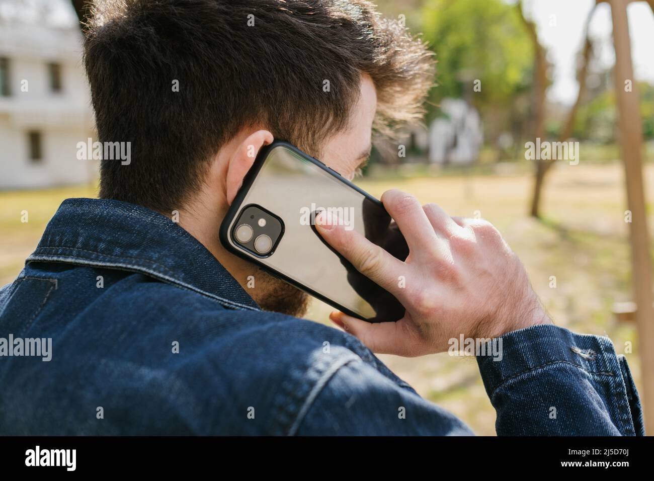 Young man wearing blue jeans jacket from behind with mobile phone ...