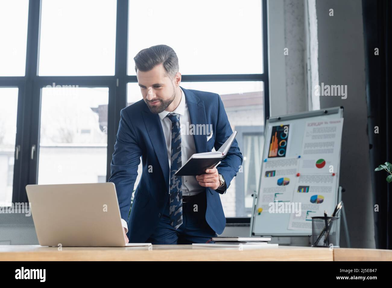 economist with notebook looking at laptop near flip chart on blurred ...