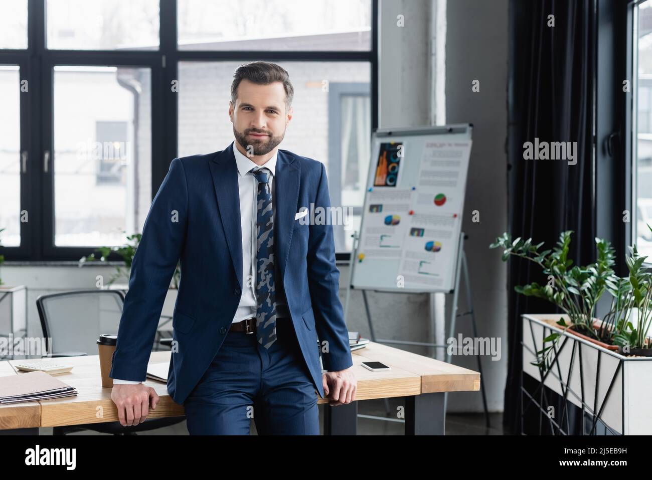positive economist standing at desk in office and looking at camera ...