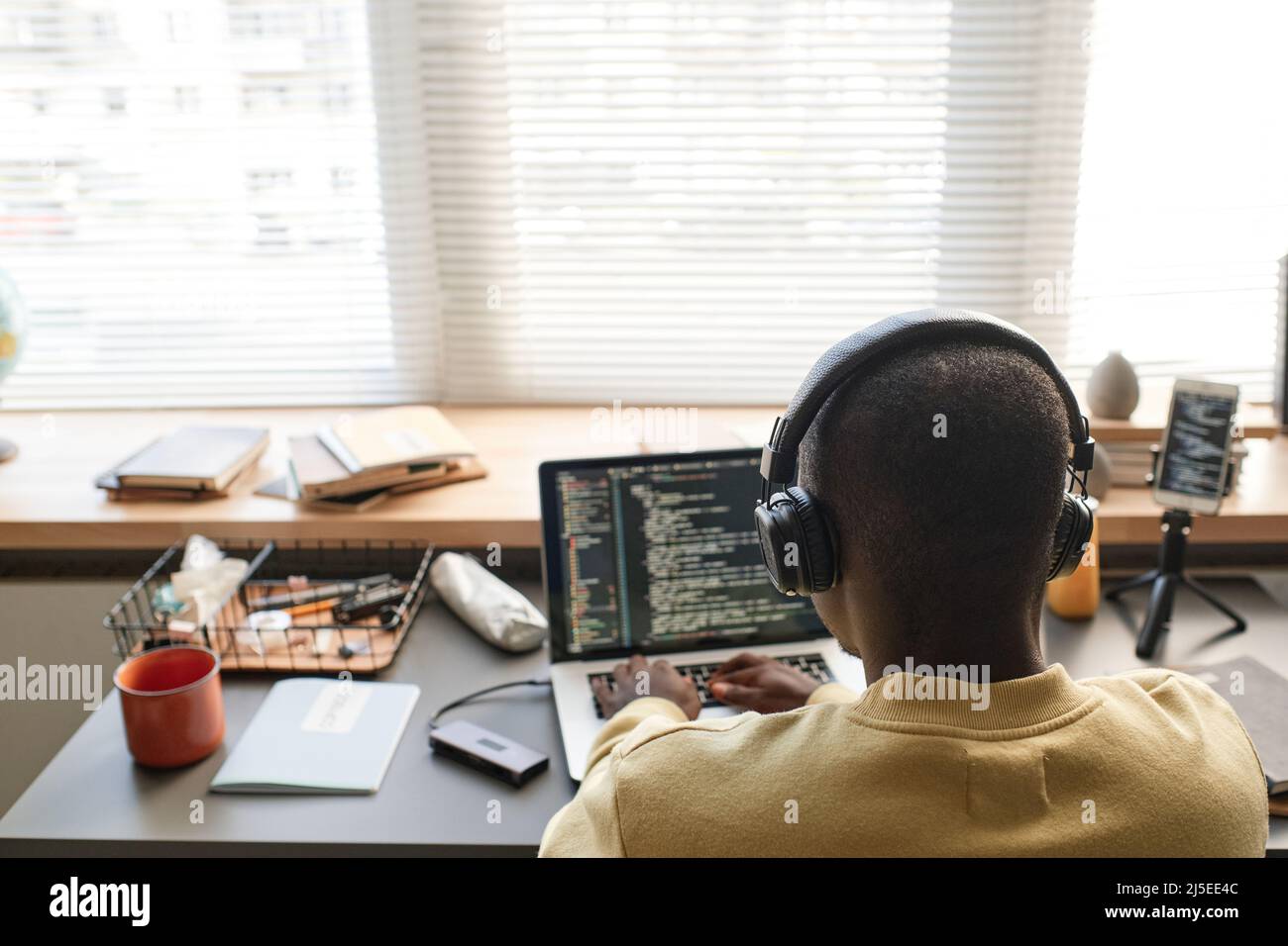 Rear view of black man in wireless headphones typing on laptop while programming code on ...