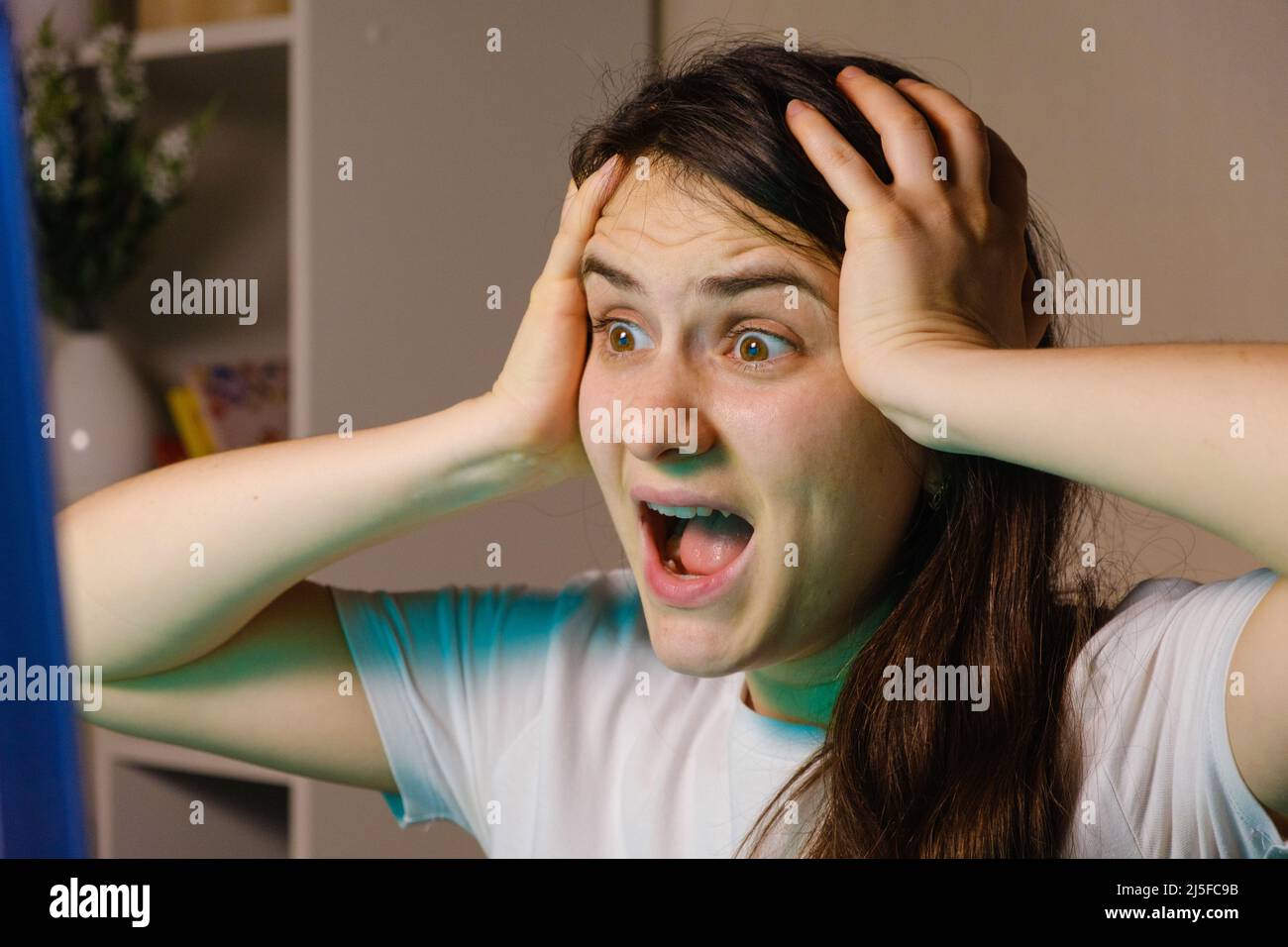 A woman screams and grabs her head with her hands while looking at a TV ...