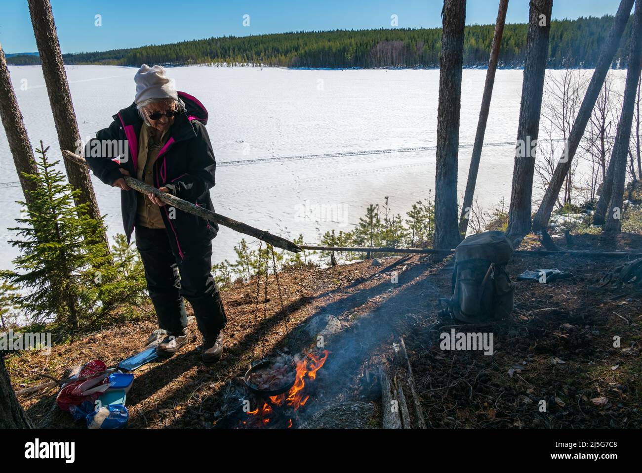 Older women holding a stick with a frying pan full of bacon over a camp ...
