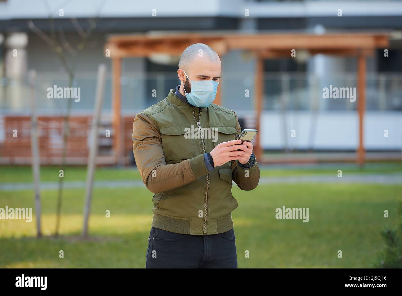 A bald man in a face mask checking his smartphone on a cozy street ...