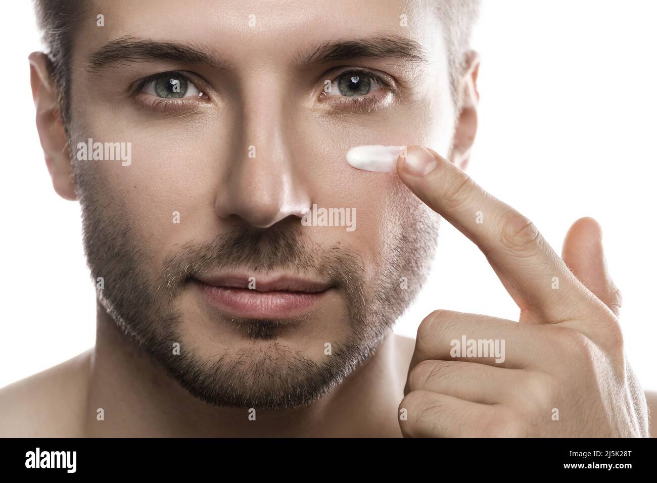 Men's beauty. Young man is applying moisturizing and anti aging cream on his face against white ...