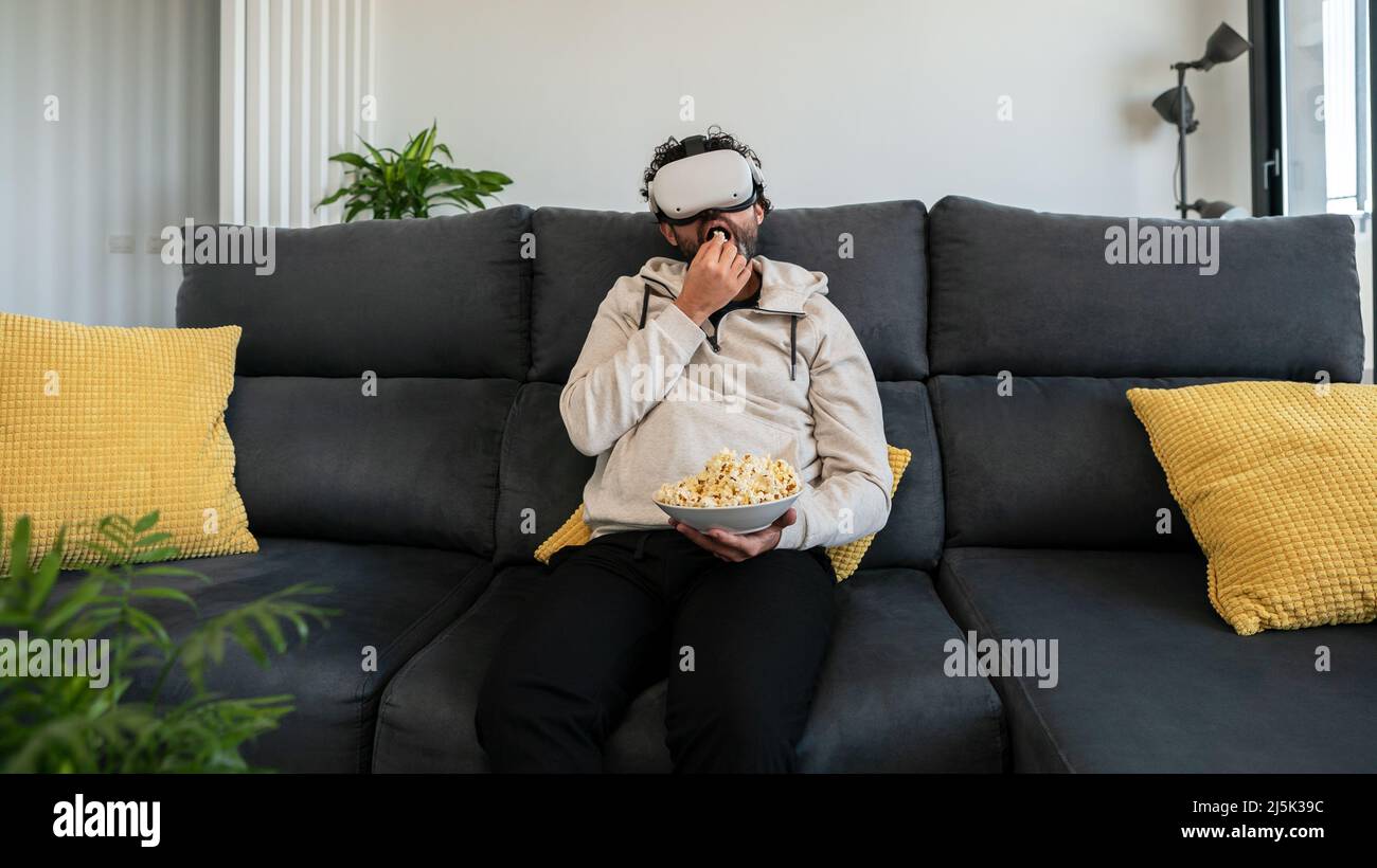 Bearded man sitting on couch and watching movie with VR glasses headset ...