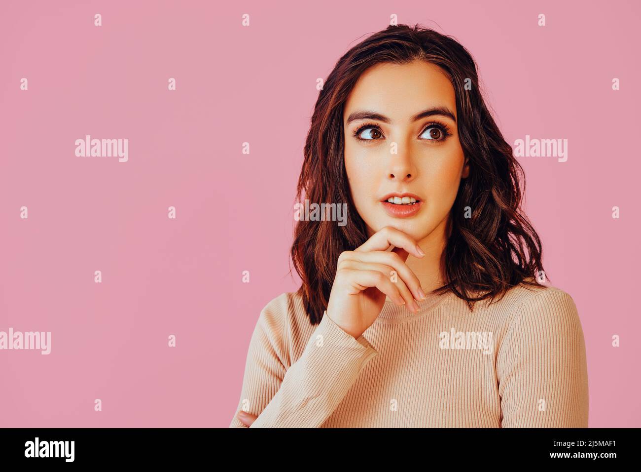 Studio portrait of thoughtful young woman with hand on chin looking up ...