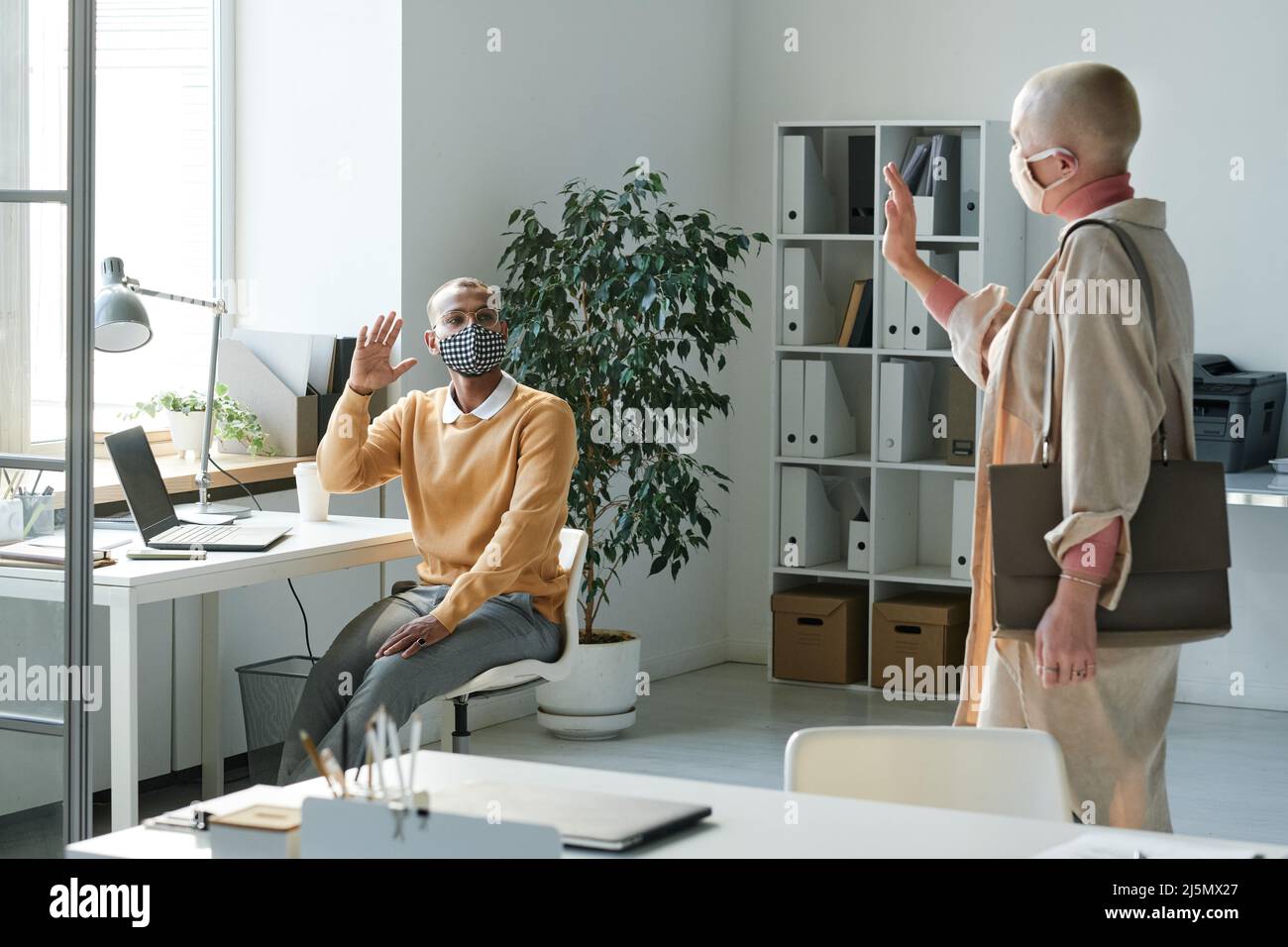 Bald woman in mask waving colleague while keeping social distance in ...