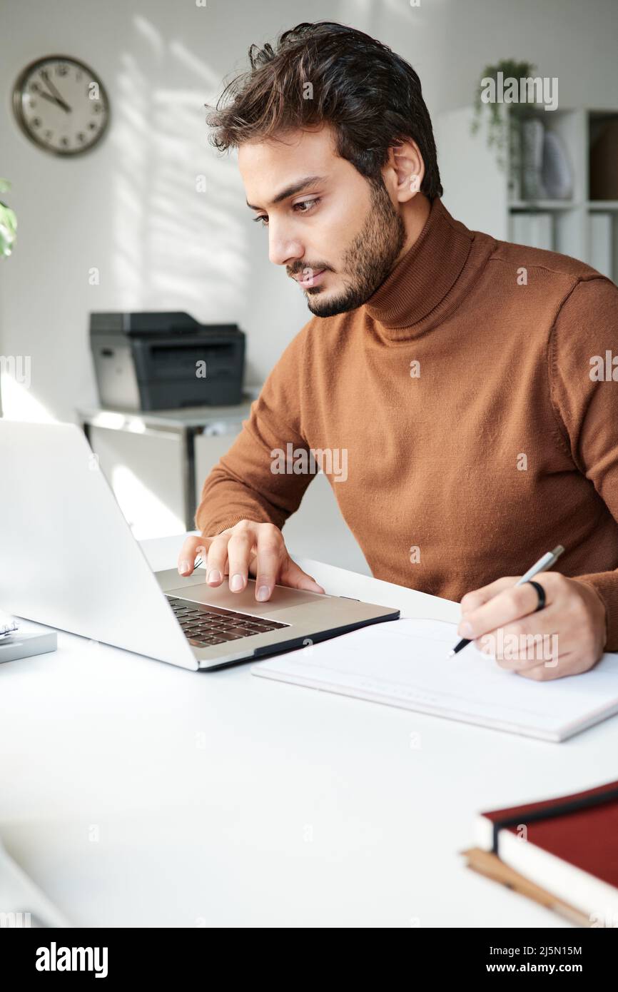 Concentrated young middle eastern man with stubble sitting at table and ...