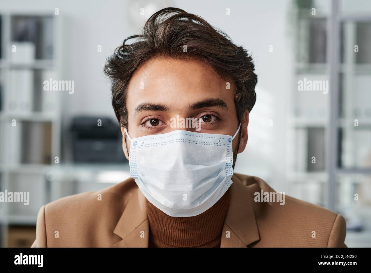 Portrait of handsome young Middle eastern manager in mask sitting in ...