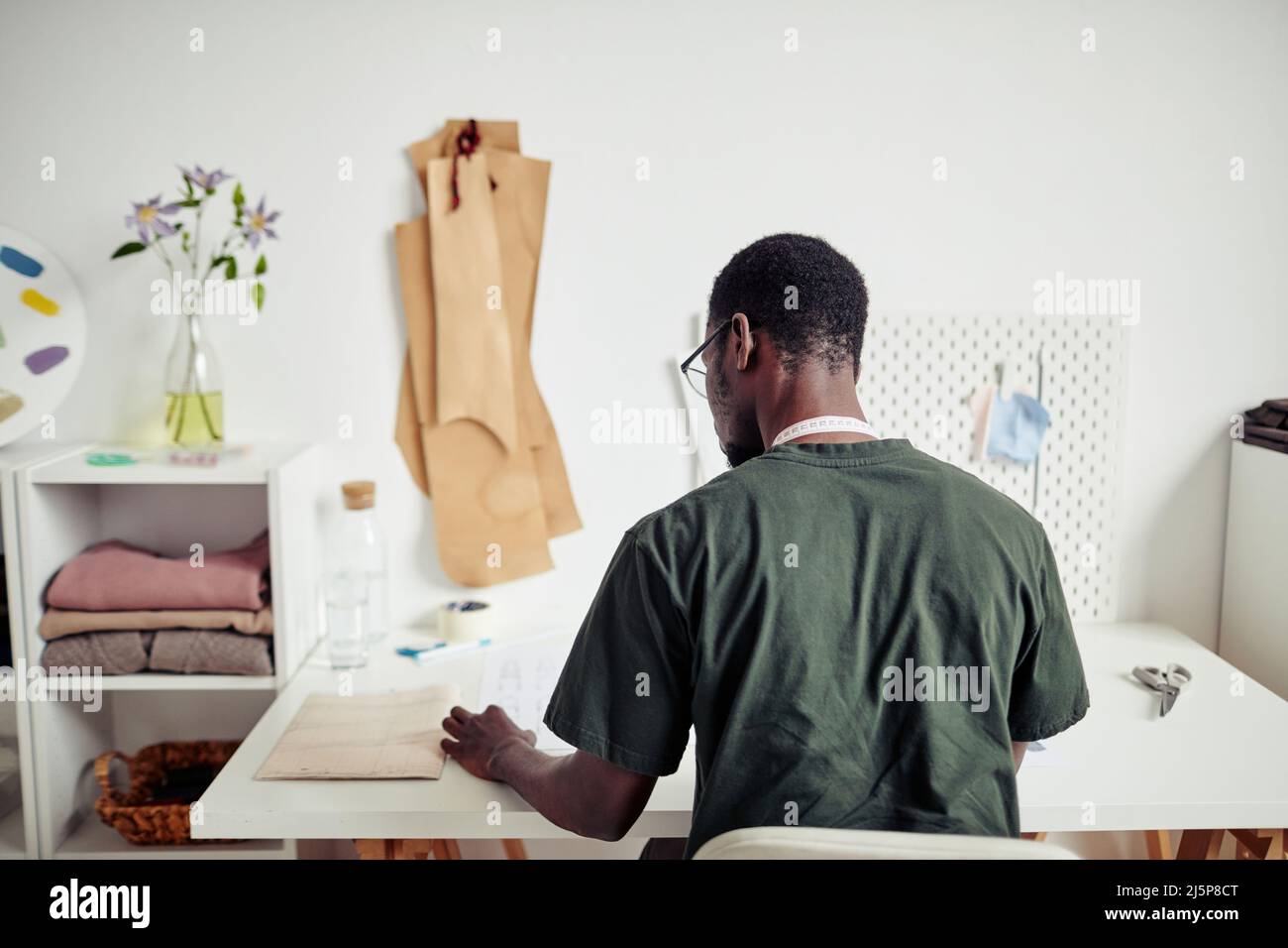 Young tailor sitting at desk in his studio and choosing sewing pattern ...