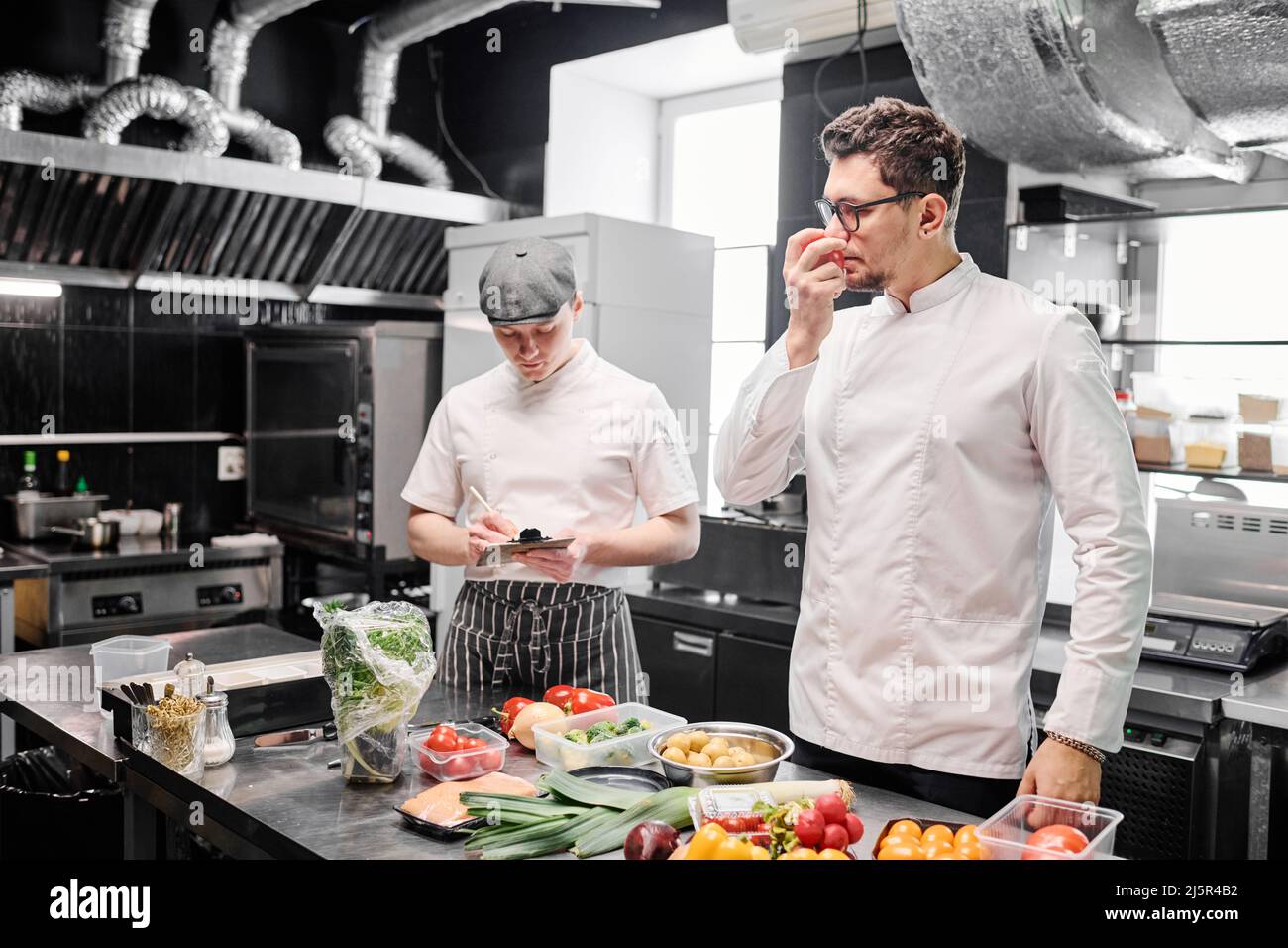 Young chef in uniform checking freshness of vegetables standing near ...