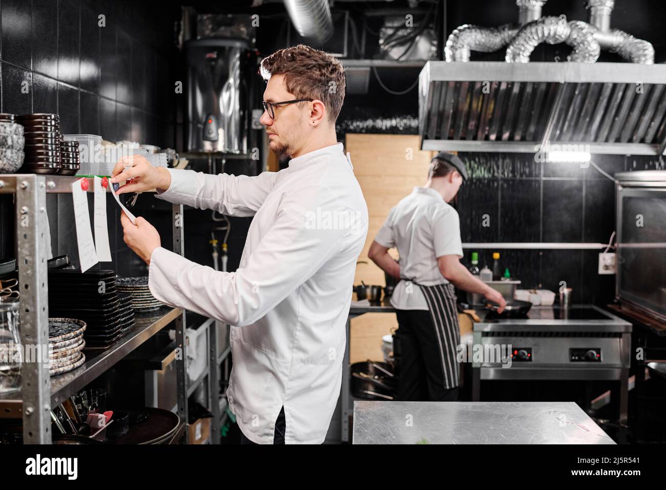 Young cook in uniform examining orders on rack with his colleague using ...