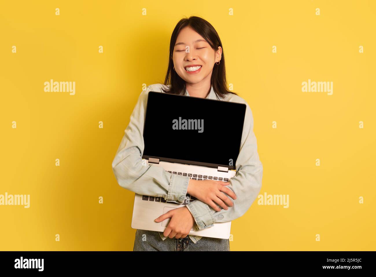 Technology lover. Cheerful young Asian woman hugging laptop computer with blank screen, mockup ...