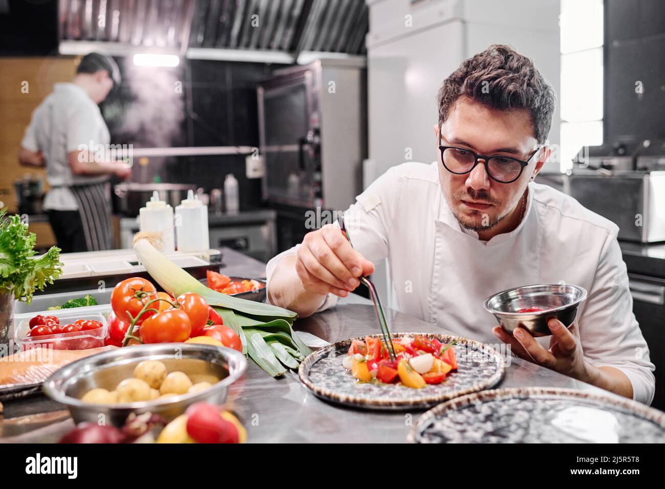 Young chef in eyeglasses decorating dish with vegetables on plate at ...