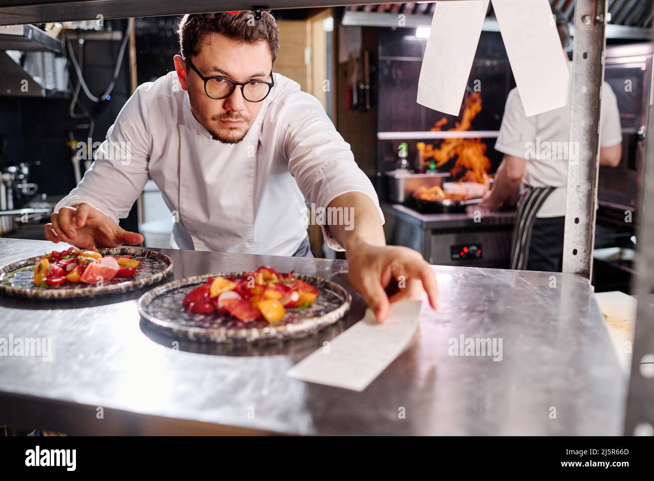 Young chef in eyeglasses putting list with order and prepared dishes on ...