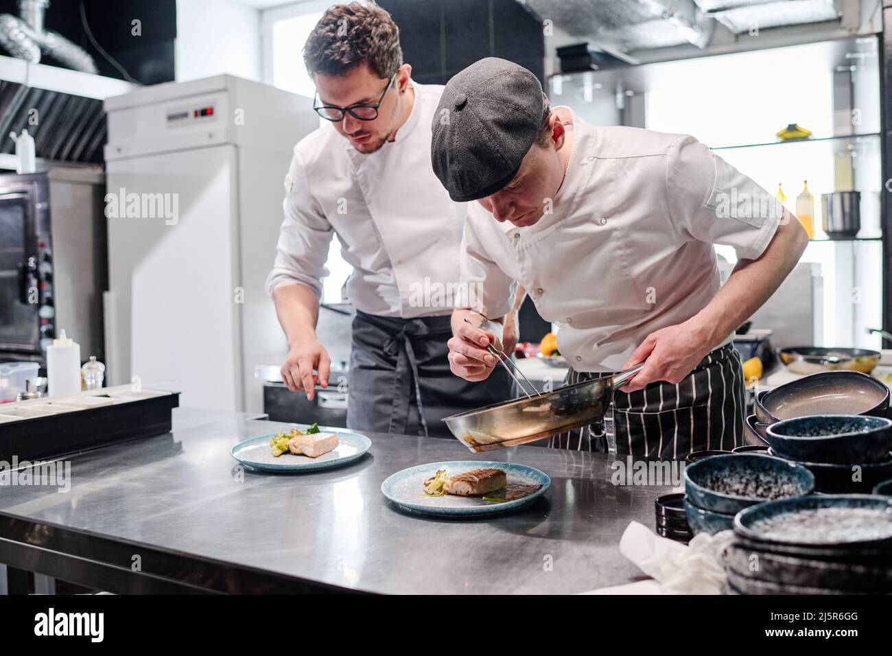 Cook in uniform putting garnish on plate from pan with chef standing ...