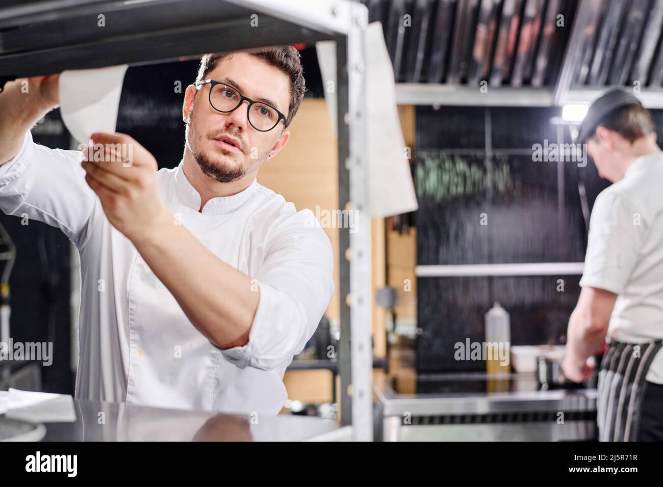 Young chef in eyeglasses taking order from counter to cook dish in ...