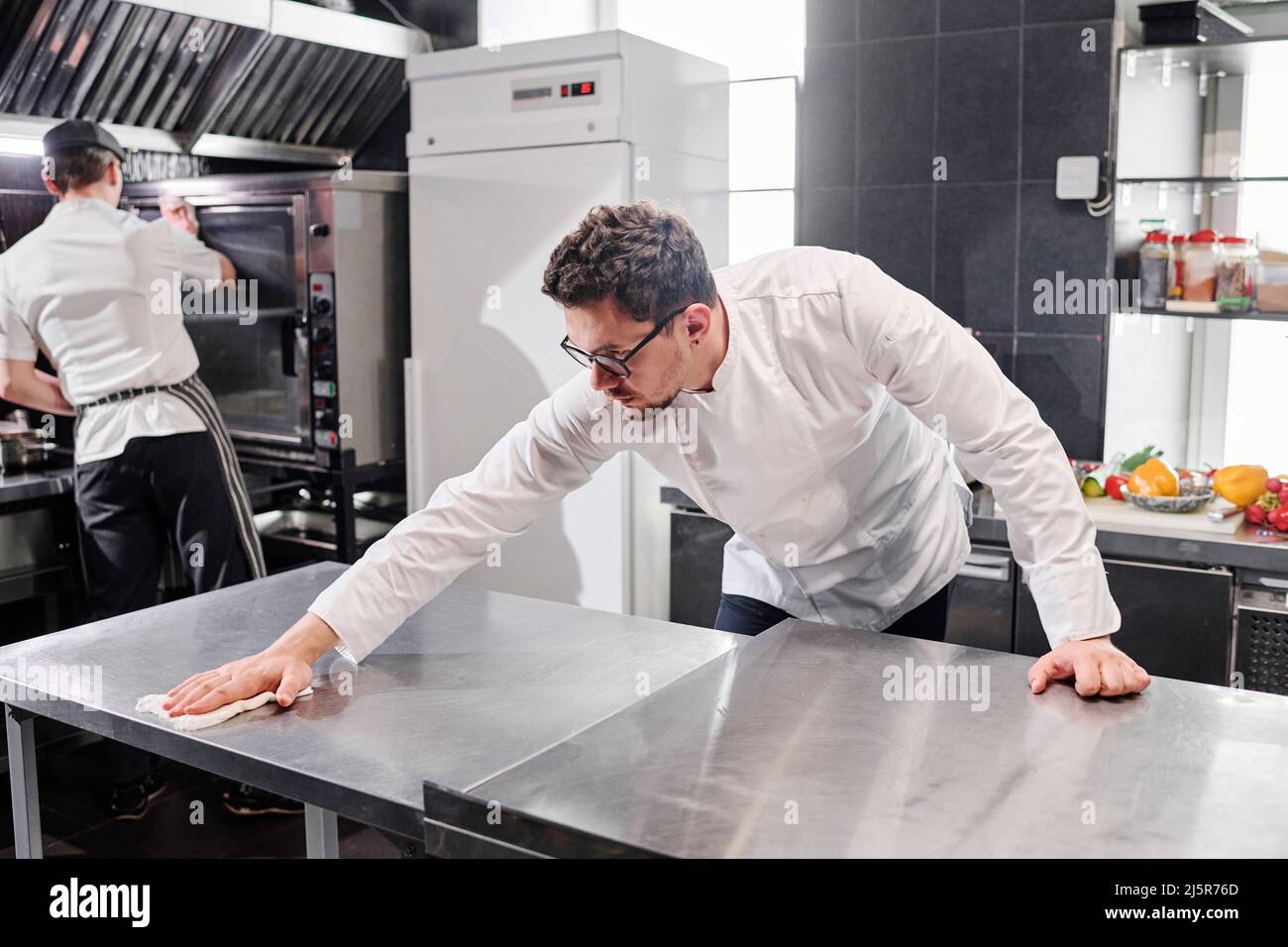 Chef in uniform wiping kitchen table after cooking with his colleague ...