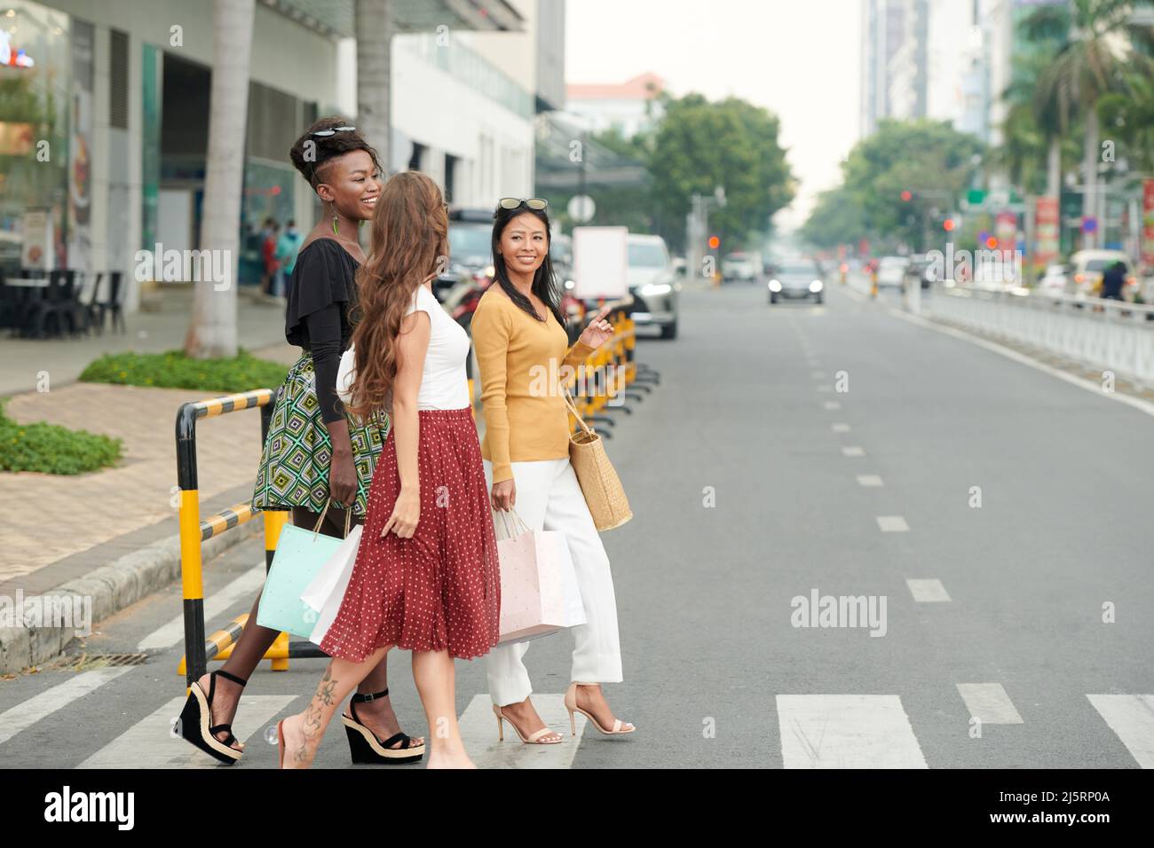 Elegant smiling young women talking when crossing road in city center ...