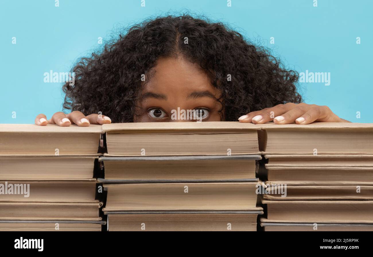 Scared African American female student hiding behind stacks of books ...
