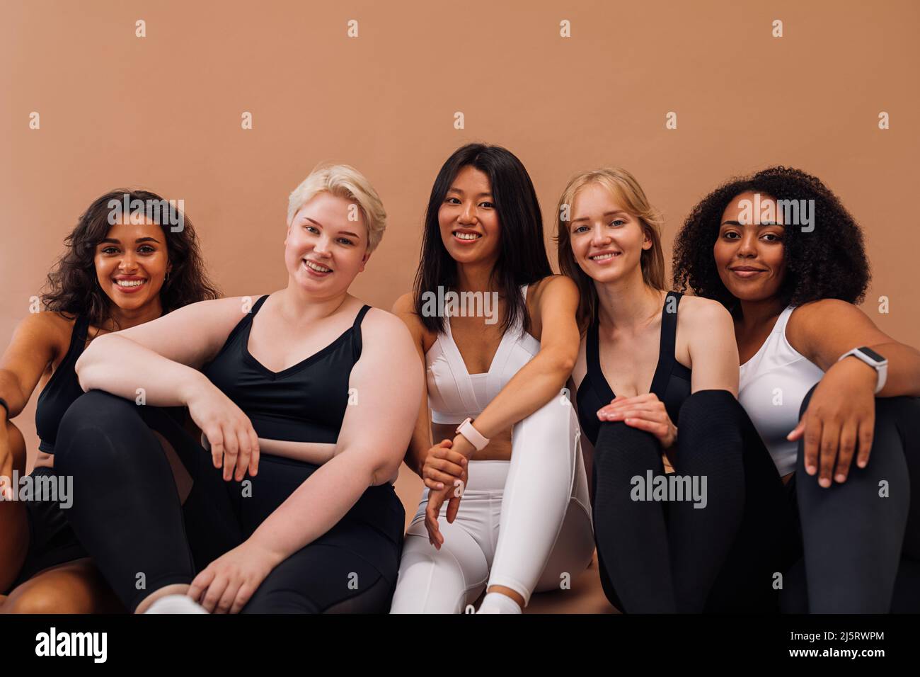 Five smiling women in fitness clothes sitting together on pastel ...
