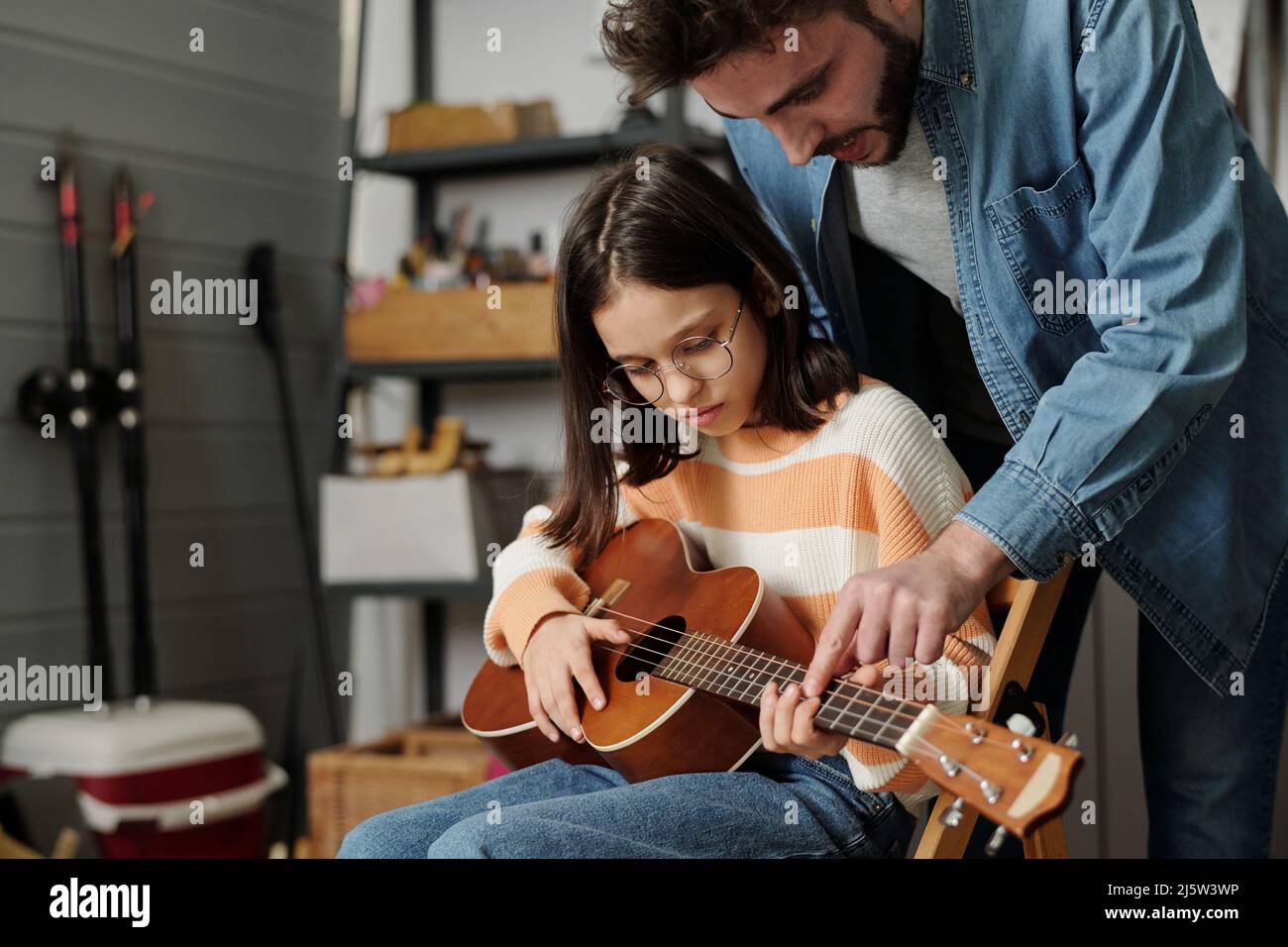 Little girl in eyeglasses learning to play guitar while young man ...