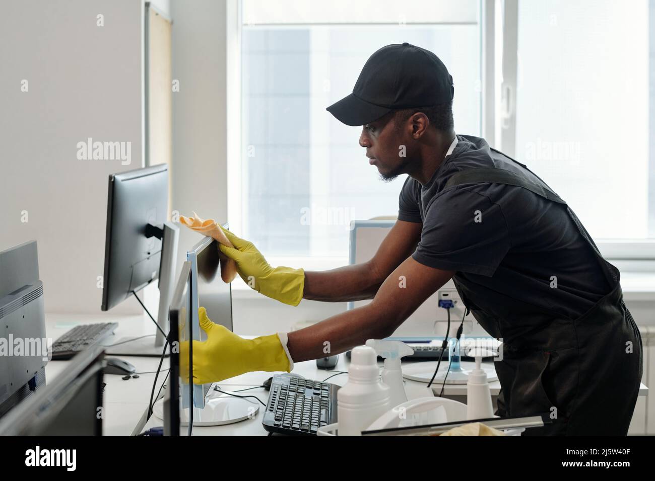 Young black man in uniform and yellow gloves wiping computer monitor ...
