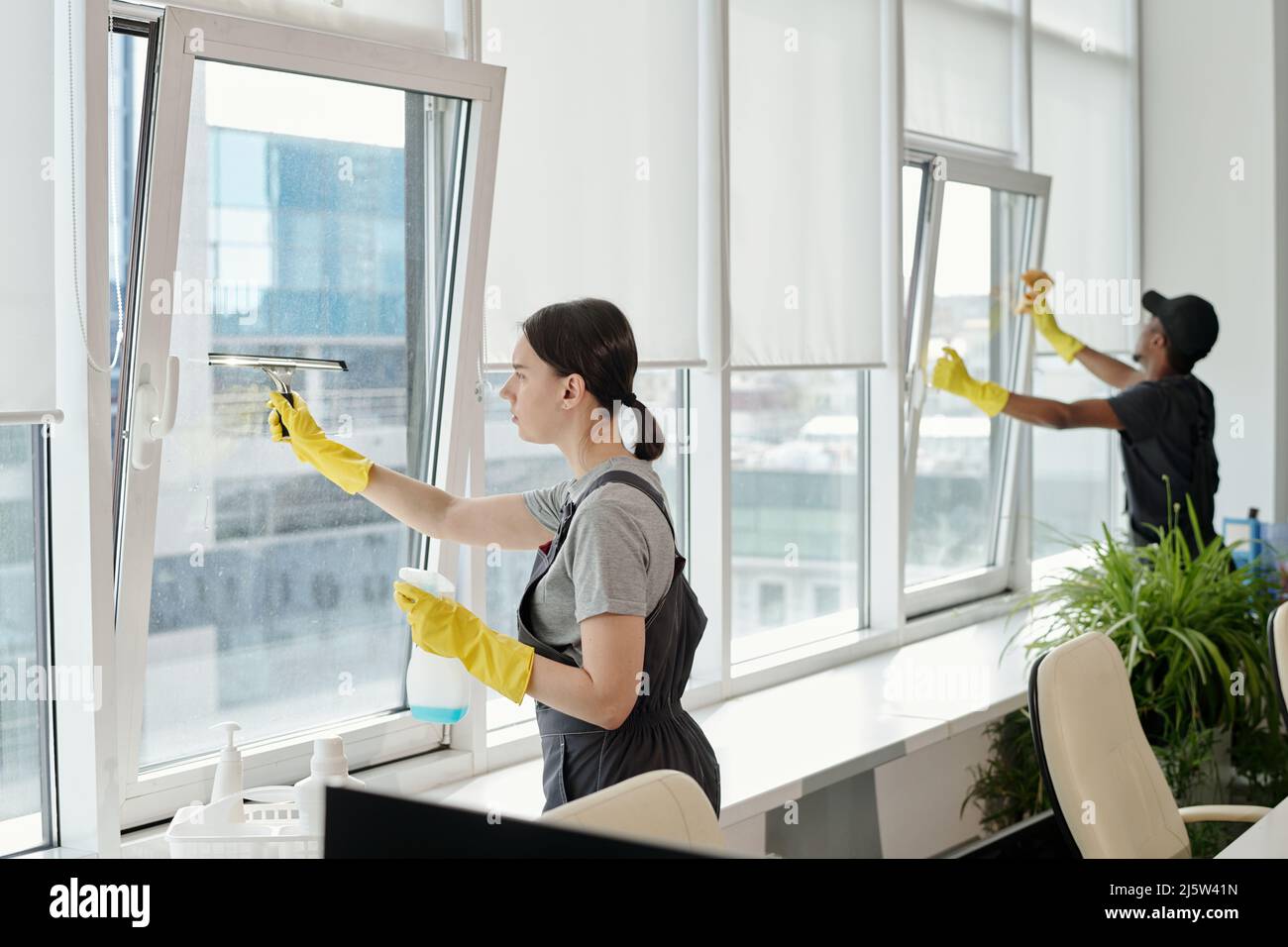 Young female cleaner in workwear washing large windows of openspace ...