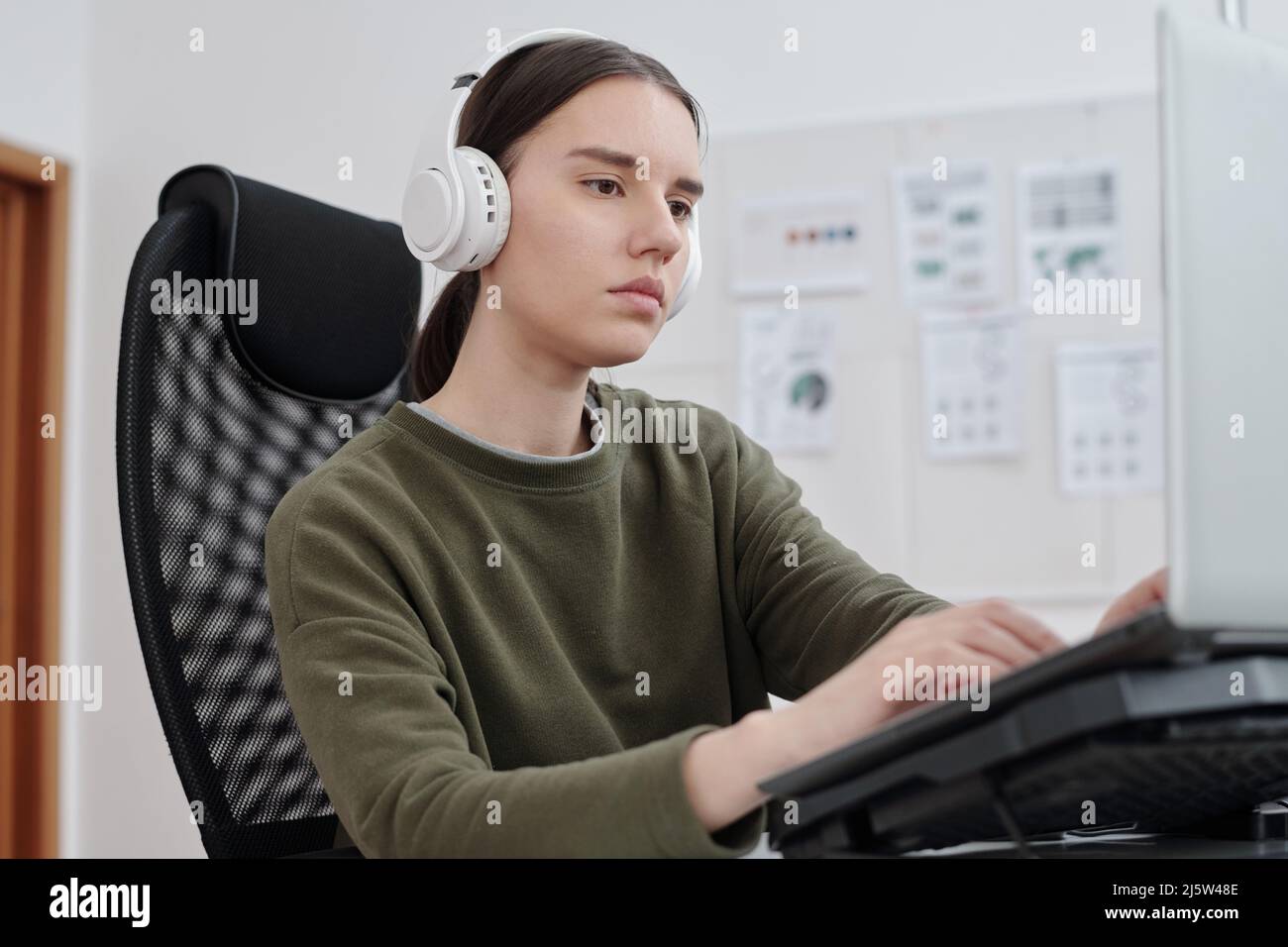 Young serious female programmer in headphones and casualwear decoding data in front of computer ...