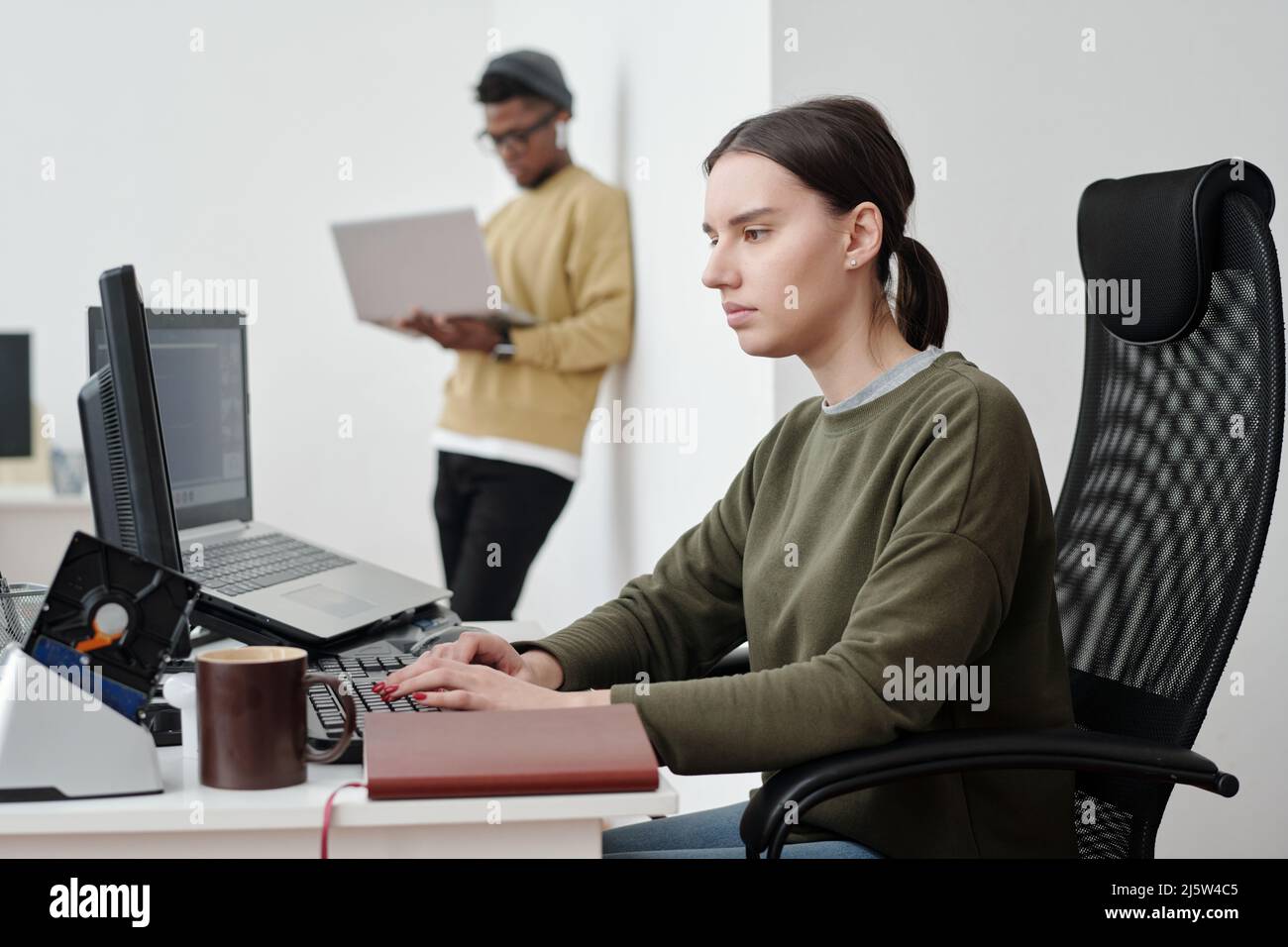 Young serious female programmer in casualwear looking at computer screen while decoding data by ...