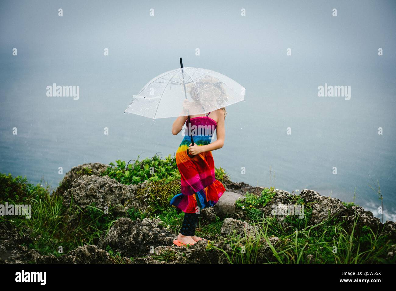 Girl plays in rain with umbrella on cliff by ocean in rainbow dress ...