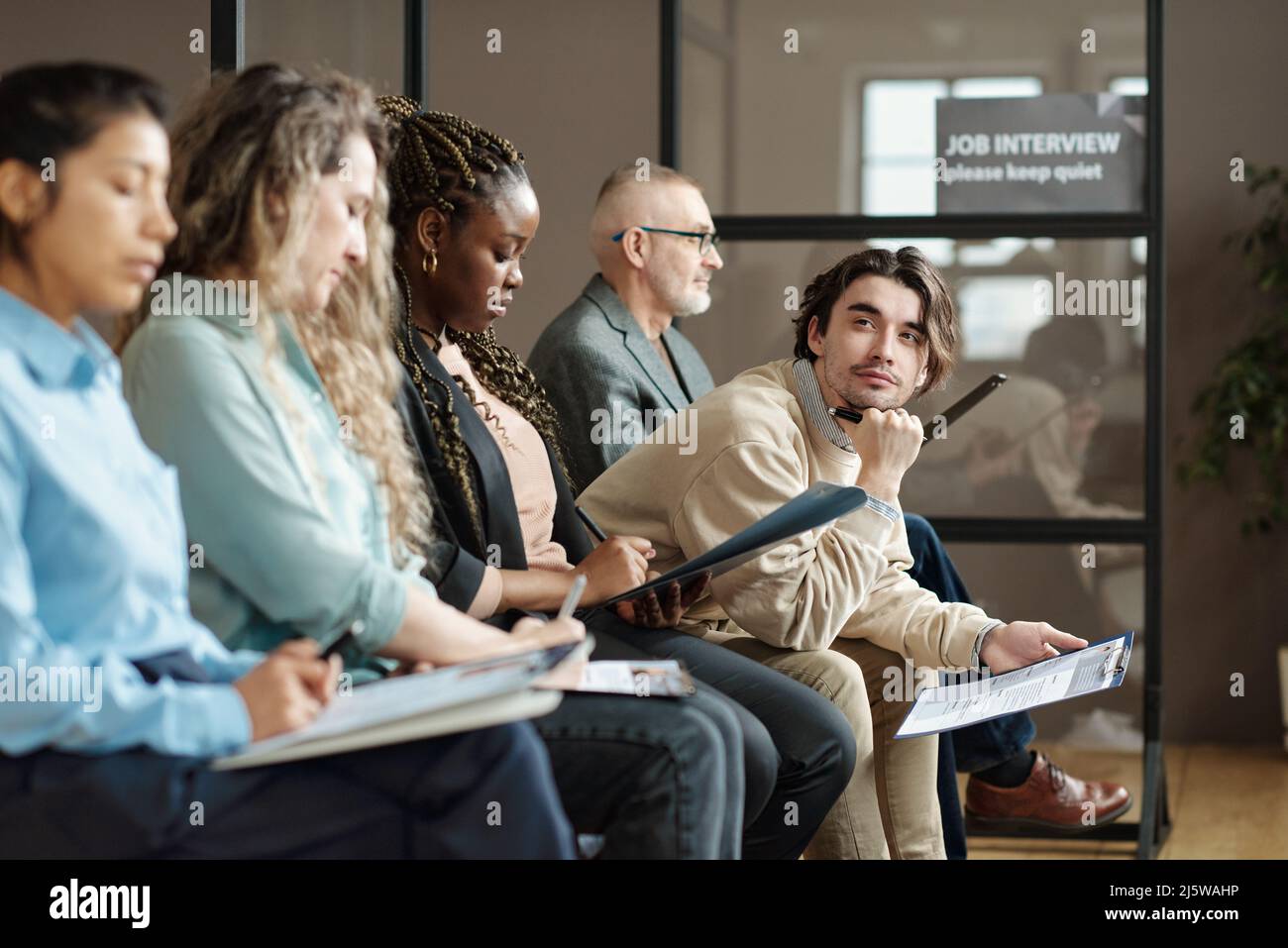 Young man sitting with resume in waiting room among other candidates ...