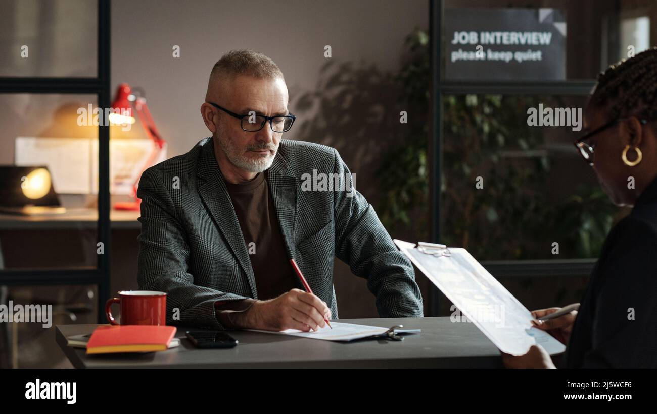 Serious mature manager in eyeglasses sitting at table and examining resume of candidate during ...