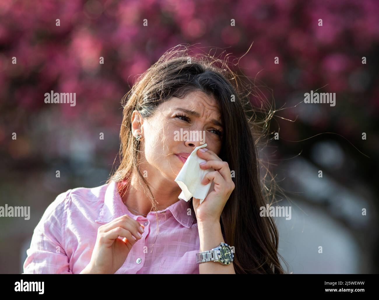 Afraid young woman wiping nose with napkin in front of blooming tree. Spring allergy attack ...