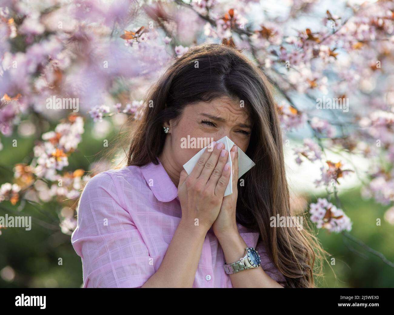 Young woman wiping nose with napkin in front of blooming tree. Spring allergy attack concept ...