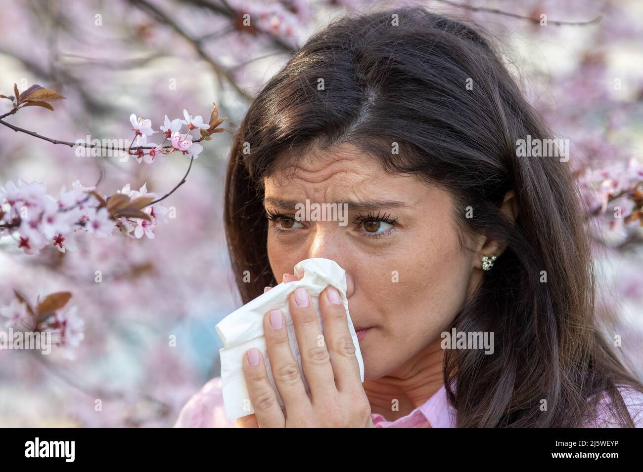 Afraid young woman wiping nose with napkin in front of blooming tree. Spring allergy attack ...