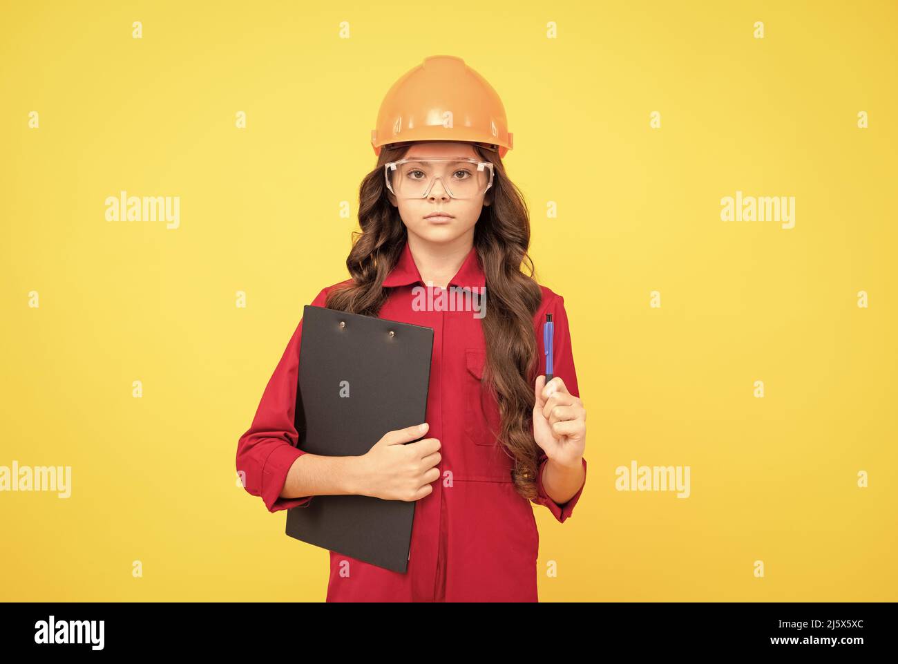 serious child girl in protective hard hat and glasses hold folder with ...