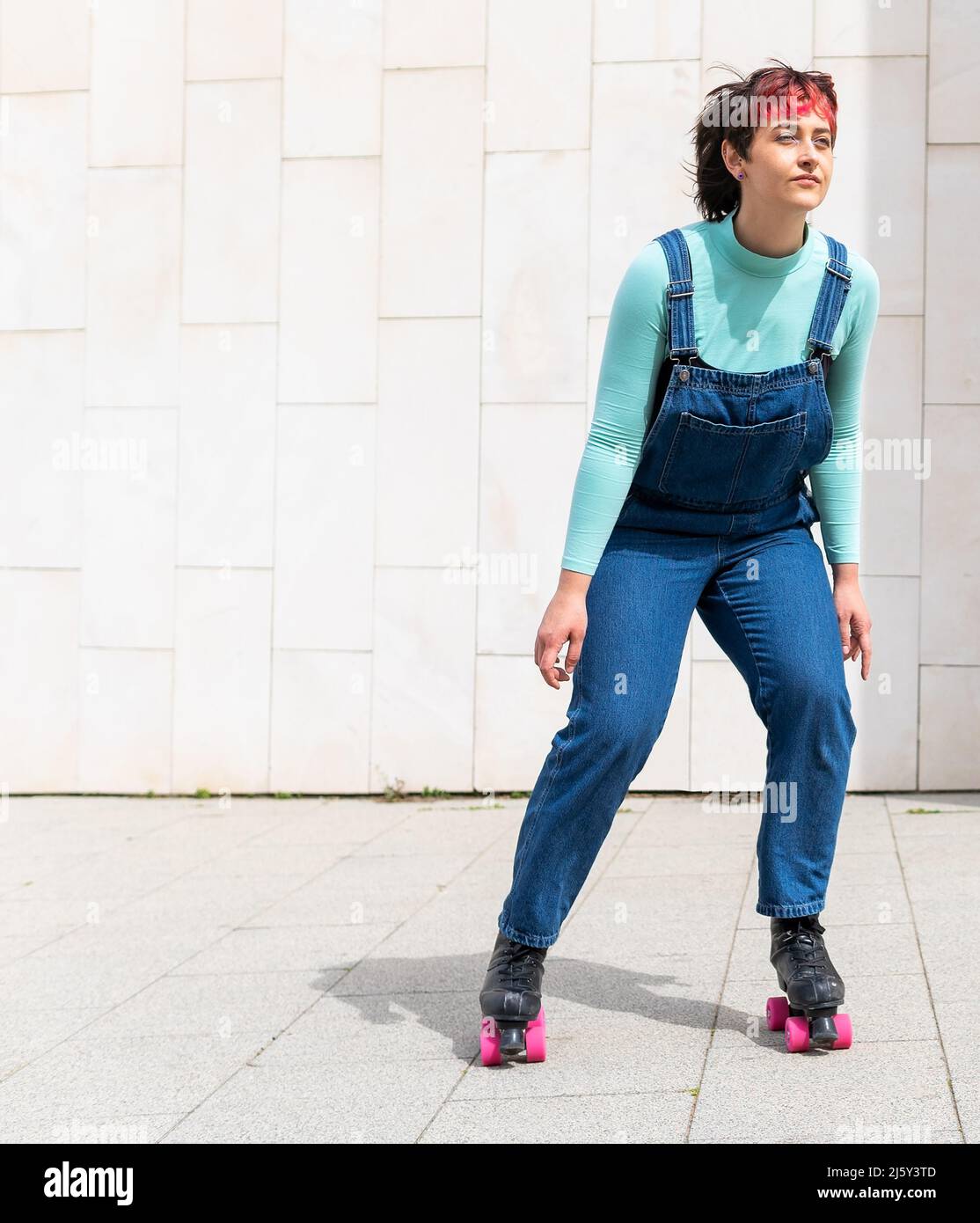 Lady with short hair in denim overall riding roller skates on pavement ...