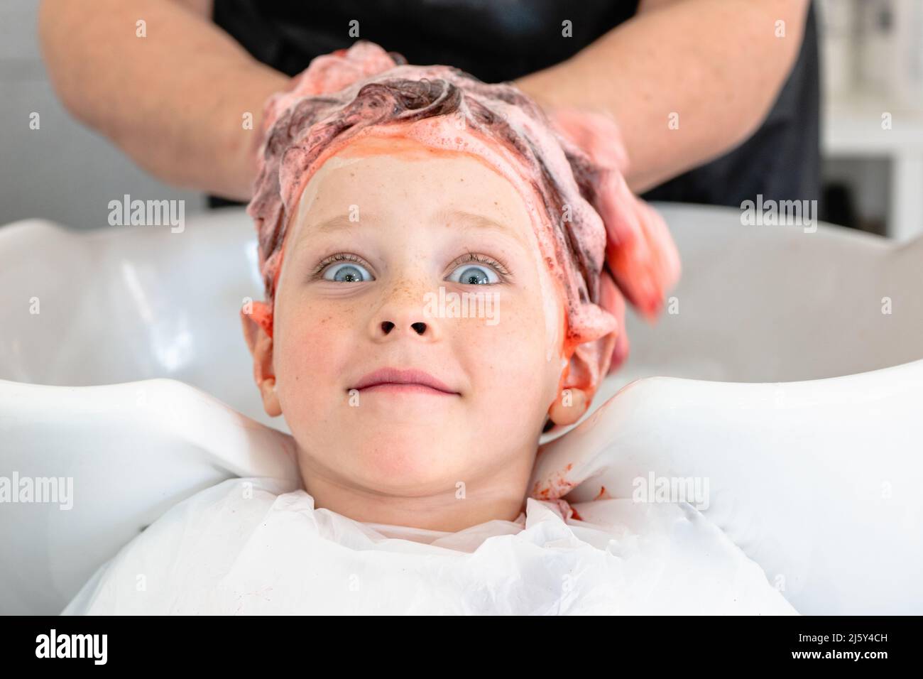 Crop unrecognizable hairstylist washing hair of amazed boy with shampoo ...