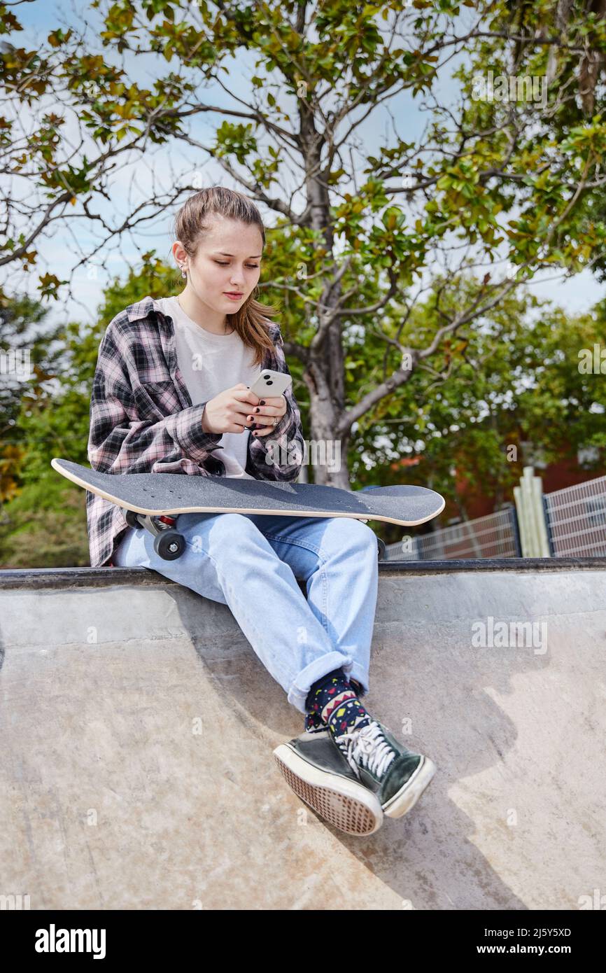 Full body of young female with black skateboard browsing on smartphone ...