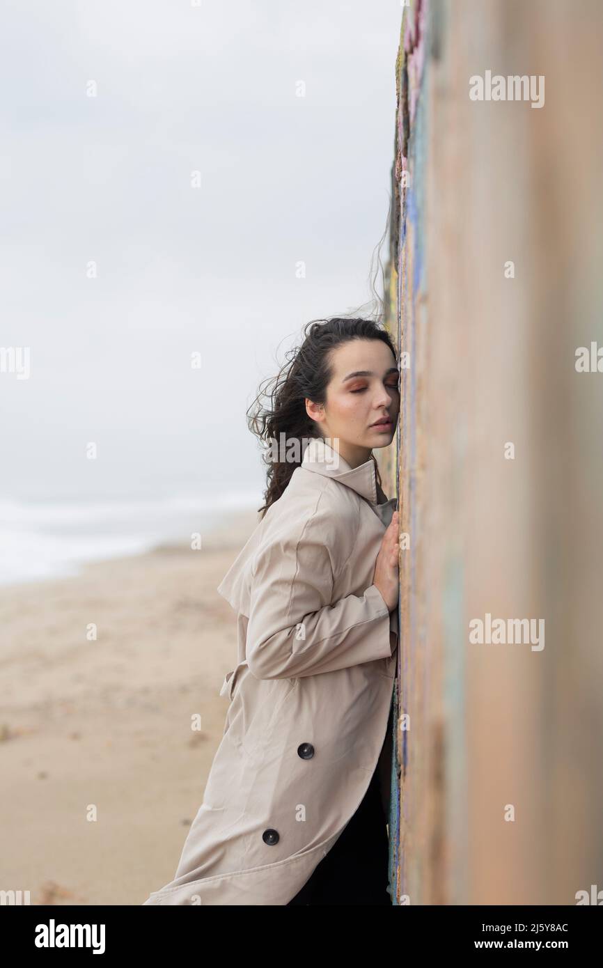 Side view of tranquil female in coat with closed eyes leaning on wall ...