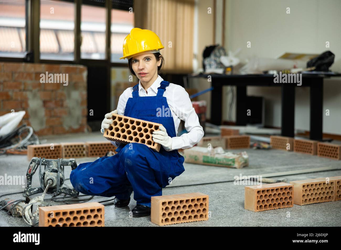 Woman builder in blue overalls lays out bricks on the wall in room ...